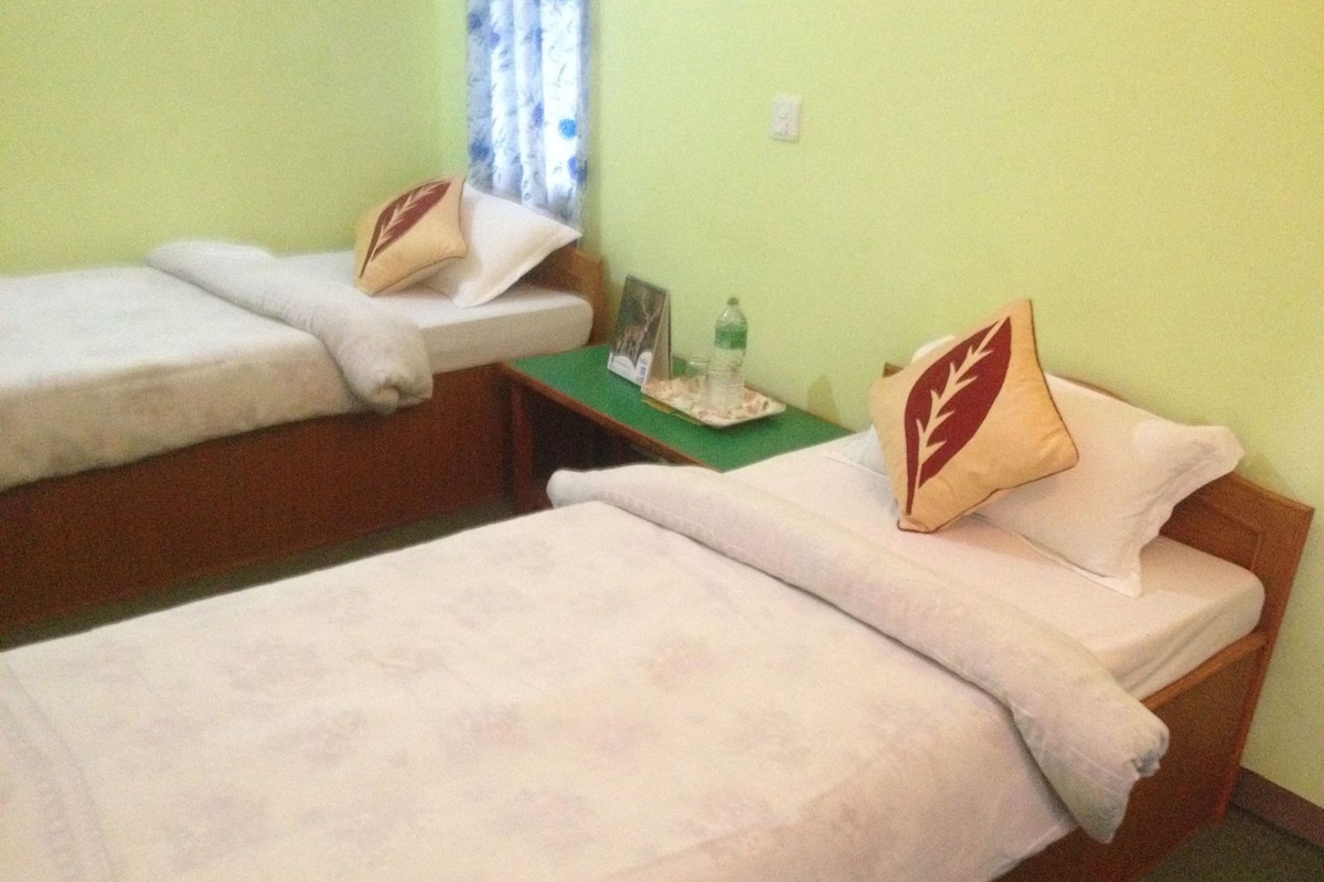 Duas camas de solteiro com almofadas combinando, uma mesinha com um copo de água e uma janela com cortinas.