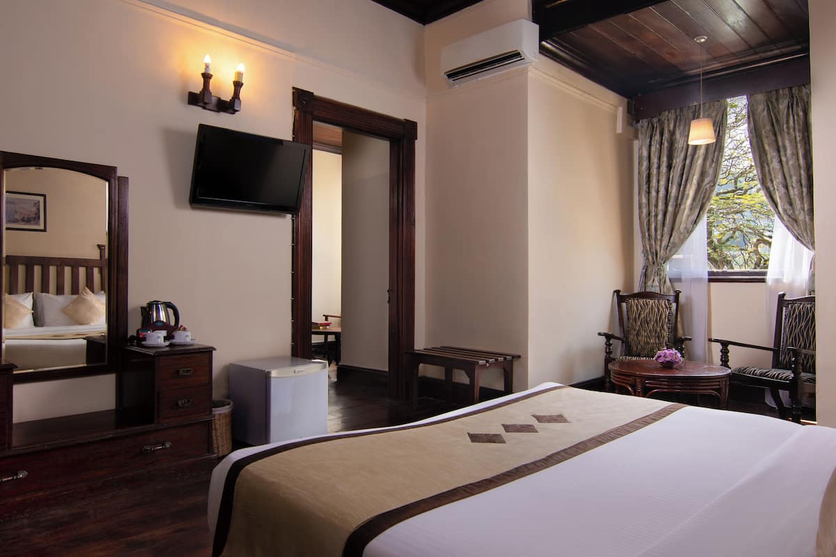 Quarto de hotel com cama, mesa de cabeceira, luminária, cadeira e televisão na parede.