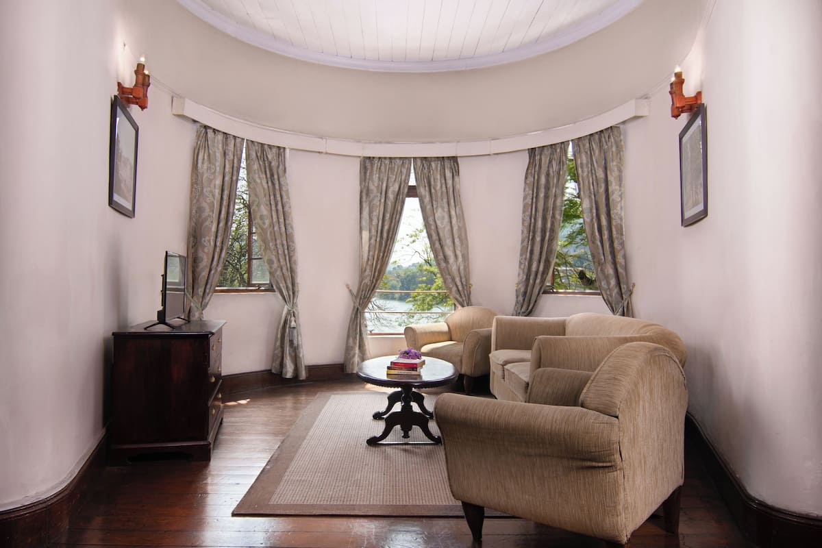 Quarto com teto curvo, uma mesa redonda com um livro e um vaso, duas poltronas bege, um armário de madeira e vista para o verde através de grandes janelas com cortinas.