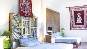 Quarto com duas camas, cabeceira de madeira, tapeçaria na parede, luminária pendente e uma pequena prateleira de madeira com objetos decorativos.