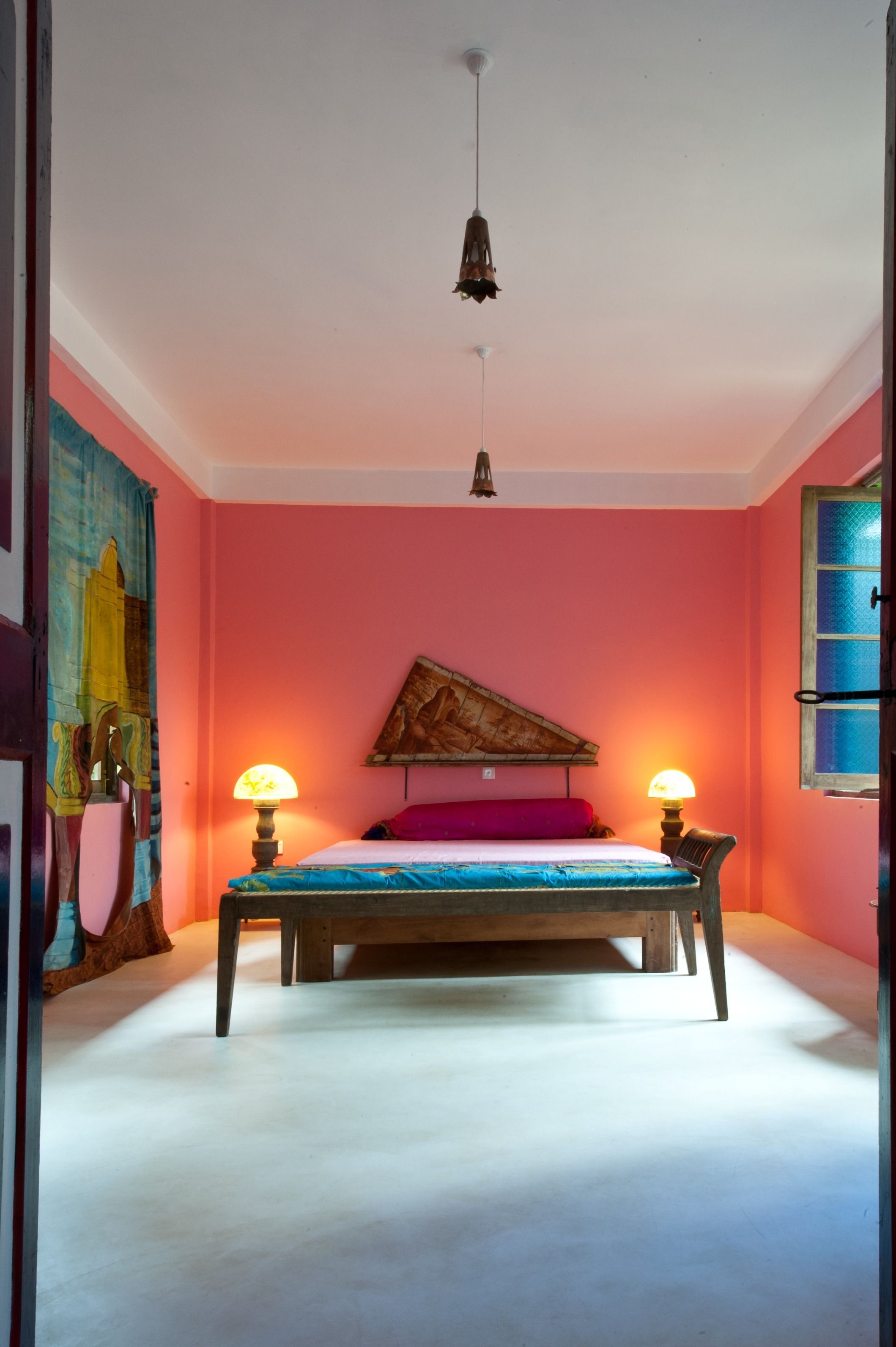 Quarto com cama, duas luminárias na parede, uma luz de teto e janela com moldura azul.