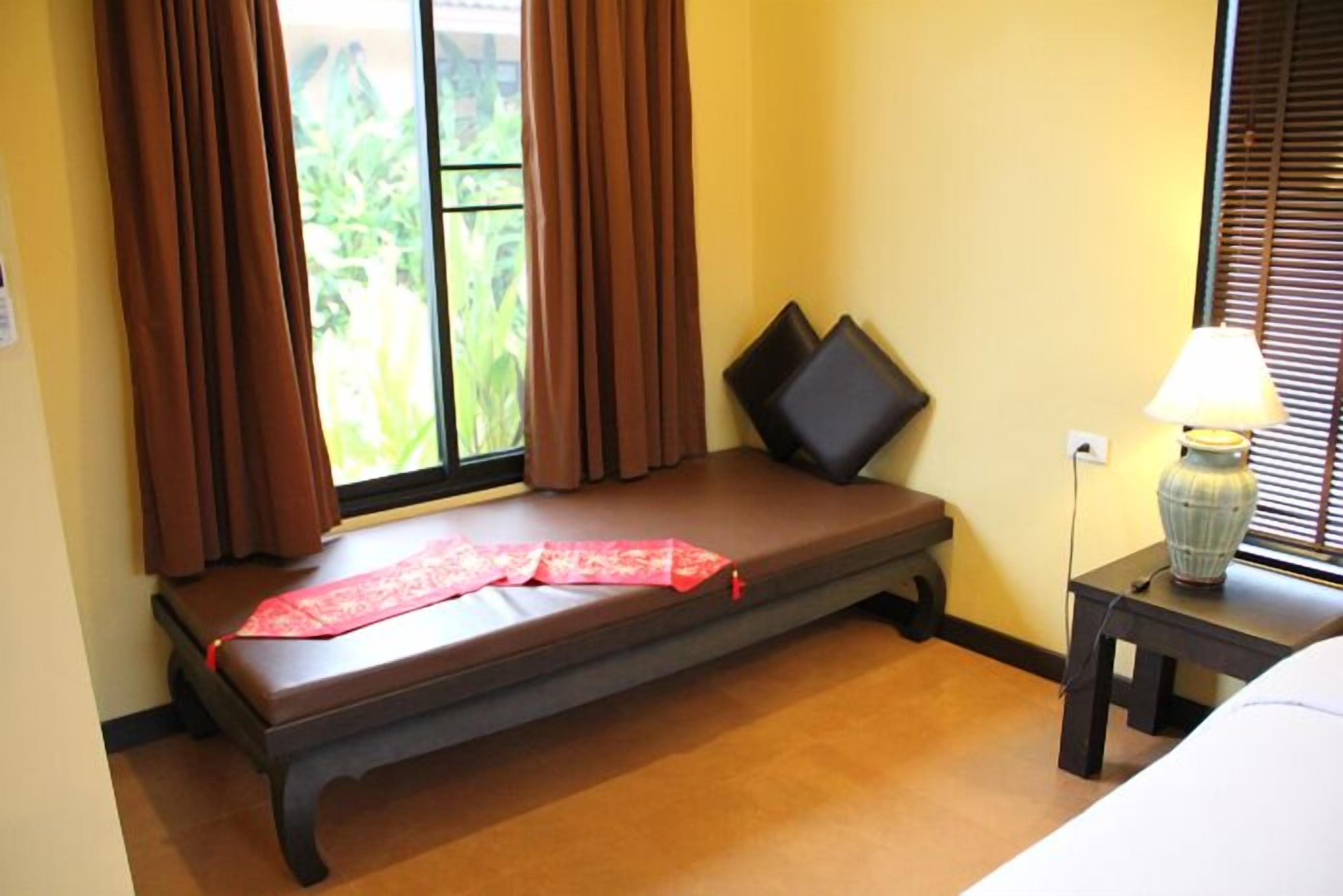 Quarto com uma cama de solteiro, um banco com tecido vermelho, uma mesinha de cabeceira com abajur e uma janela com cortinas.