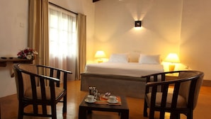 Quarto de hotel com cama, duas cadeiras, uma mesinha com chávenas e um tabuleiro, e uma mesa de cabeceira com uma lâmpada.
