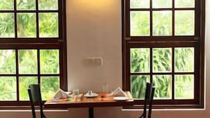 Quarto com uma mesa e duas cadeiras, amplas janelas com vista para área verde e luminária na parede.