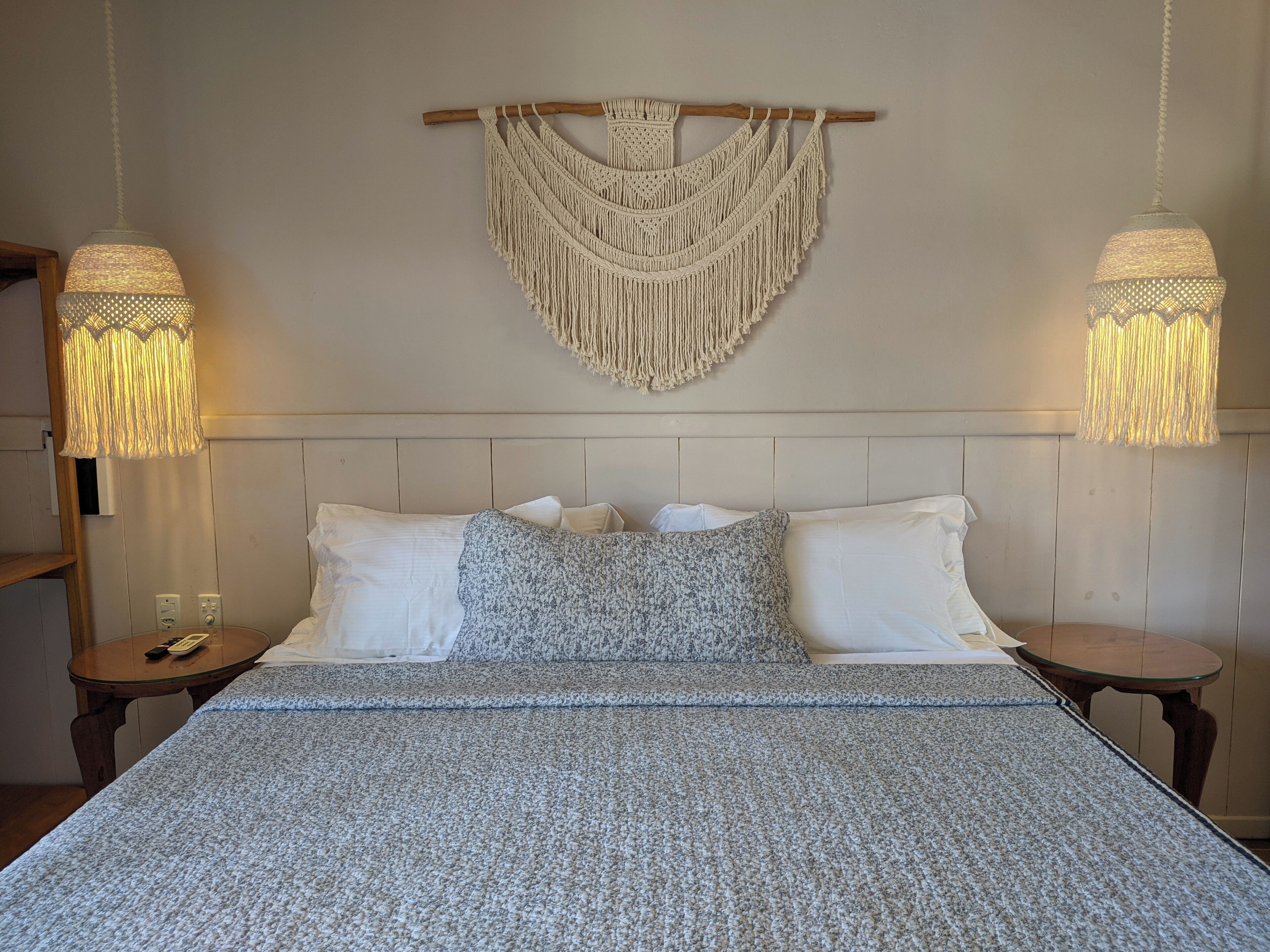 Uma cama com colcha estampada, duas mesas de cabeceira e peças de macramé na parede.