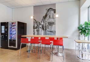 Uma área de bar moderna com um mural de uma cena urbana, uma máquina de venda automática e banquetas vermelhas.