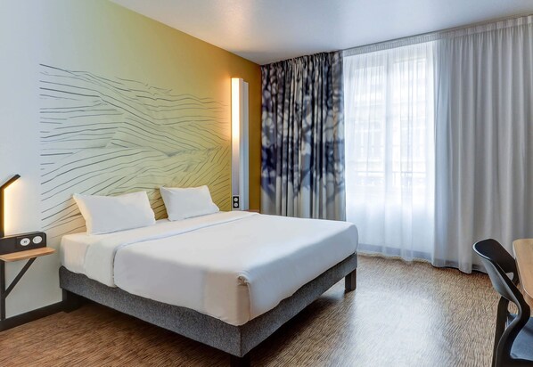 Quarto de hotel moderno com uma cama grande, uma mesa de cabeceira com abajur, uma obra de arte na parede e vista para o exterior através de cortinas translúcidas.