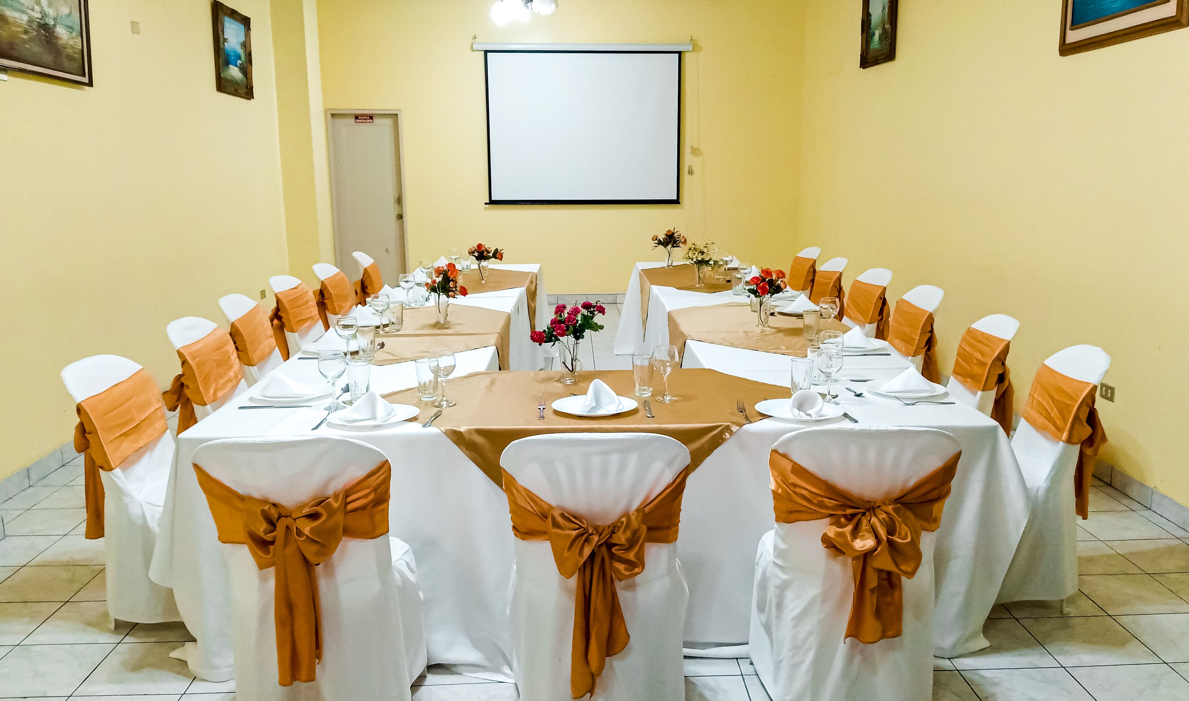 Uma sala de conferências com uma mesa comprida preparada para uma reunião, cadeiras brancas com fitas laranja e uma tela de projetor na parede.