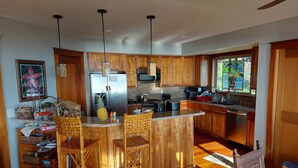 Uma cozinha moderna com armários de madeira, uma ilha central com banquetas, uma geladeira de aço inoxidável e uma janela que ilumina naturalmente.