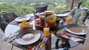 Uma mesa de jantar posta com pratos, talheres e diversos alimentos, rodeada por cadeiras com almofadas estampadas, em um terraço com vista panorâmica.