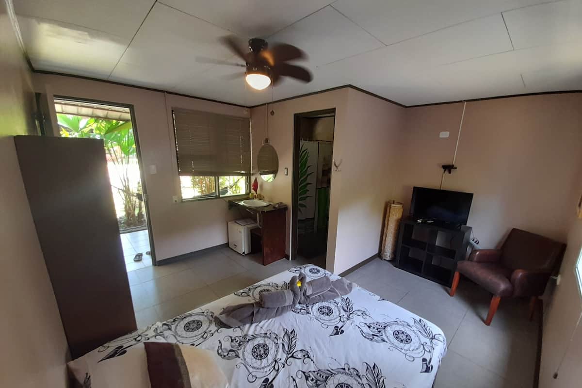 Quarto com cama, televisão, cadeira, ventoinha no teto, janela e porta para um banheiro.