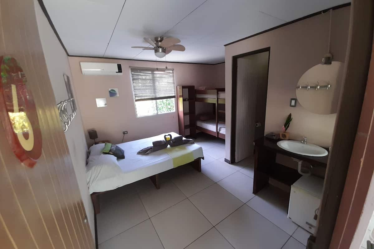 Quarto com uma cama de solteiro, ventilador de teto, janela com persianas, beliche, banheiro com pia e espelho, e porta que dá para outro cômodo.