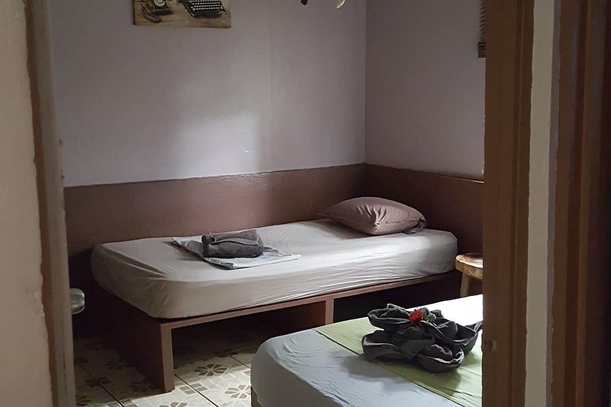 Quarto pequeno e com pouca luz, com duas camas, um ventilador de teto e um quadro pendurado na parede.