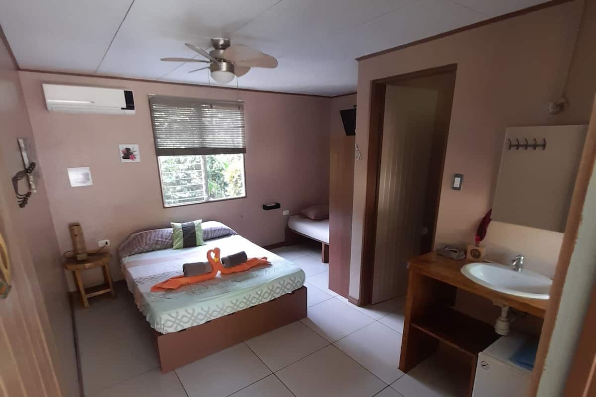 Quarto com cama, ventilador de teto, ar condicionado, janela com persianas, mesa de cabeceira e um banquinho pequeno.