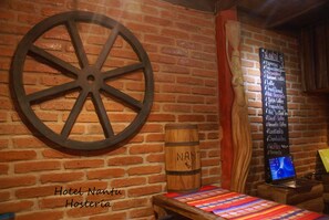 Paredes de tijolos com uma grande roda de madeira, um barril de madeira com a etiqueta "NANTU" e um quadro de menu listando diversos itens de café e comida.
