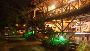 Um edifício rústico com telhado de colmo, vigas de madeira e interior em tijolo. Há plantas iluminadas em tons de verde e um caminho que leva à entrada.