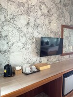 Quarto de hotel com parede de mármore, televisão de tela plana acima de um balcão de madeira, cafeteira, xícaras e um livro.