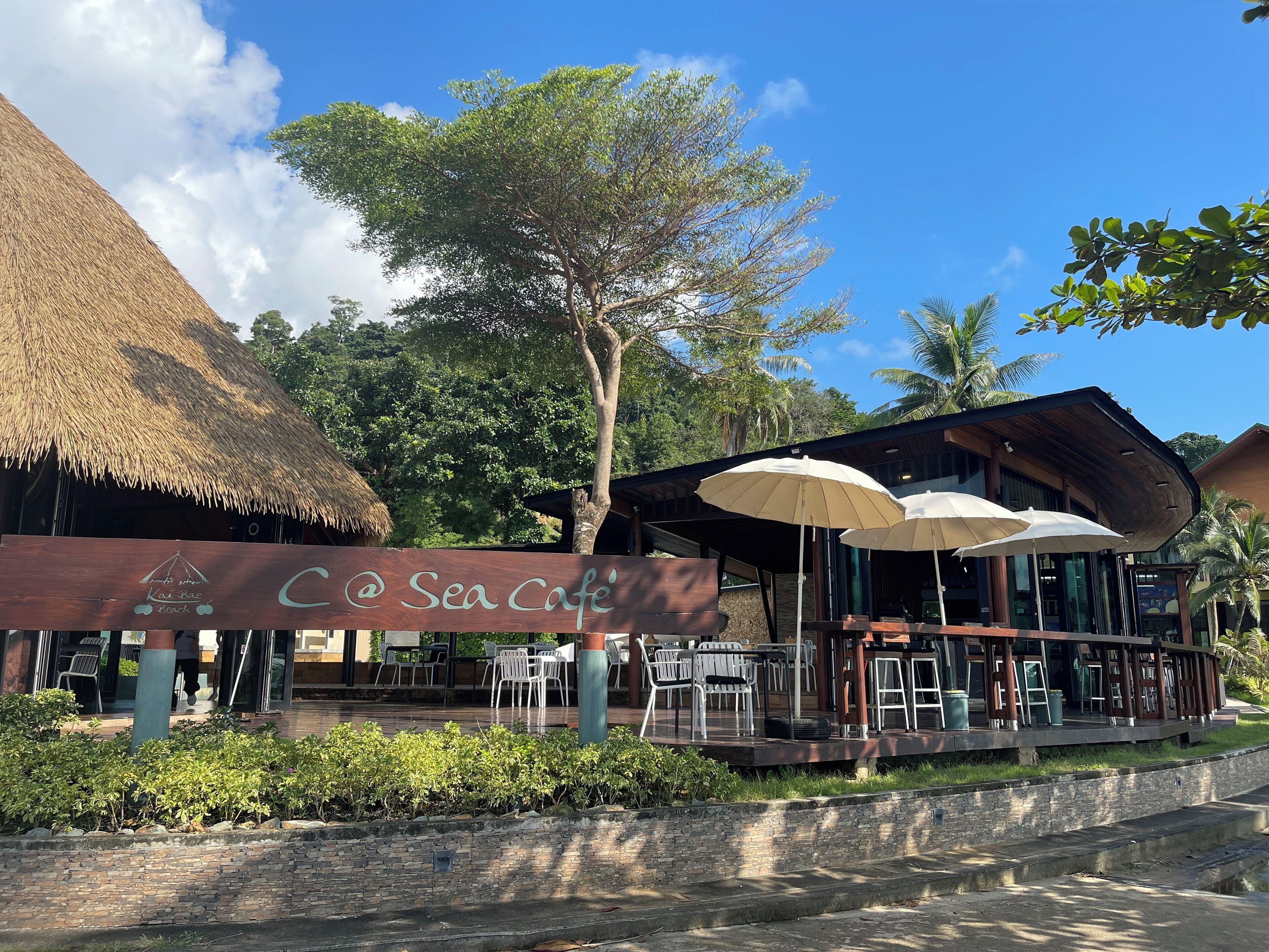 Um café à beira-mar com mesas ao ar livre, estrutura com telhado de palha e vegetação exuberante.
