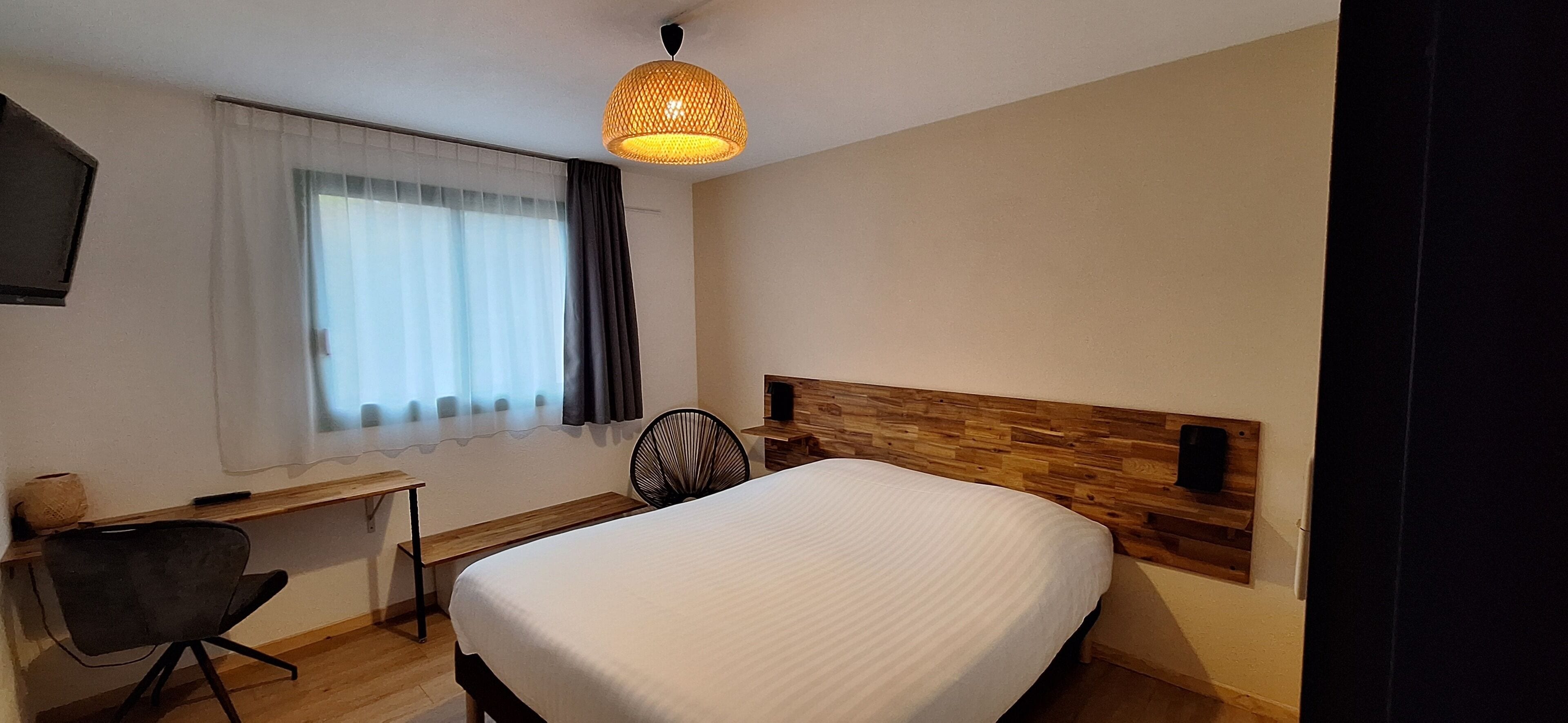 Quarto de hotel com cama, escrivaninha, cadeira, televisão e janela com cortinas.