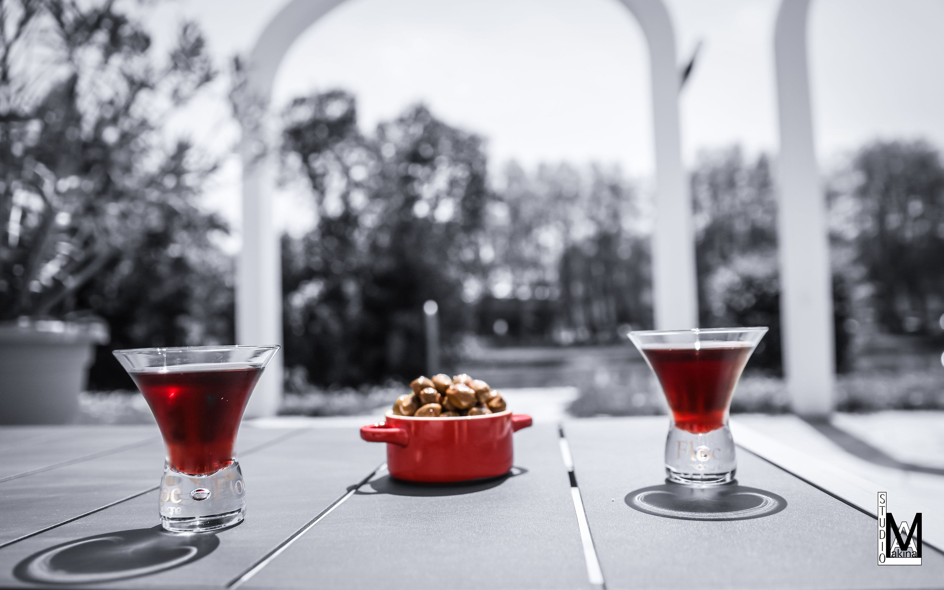 Dois copos de martini com líquido vermelho sobre uma mesa, um pote vermelho com nozes e o logotipo 'Studio M' em um apoio para copos.