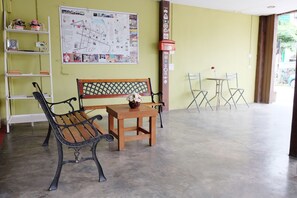 Um quarto com um banco de madeira, uma pequena mesa e duas cadeiras. Há um mural na parede com papéis diversos e um mapa.