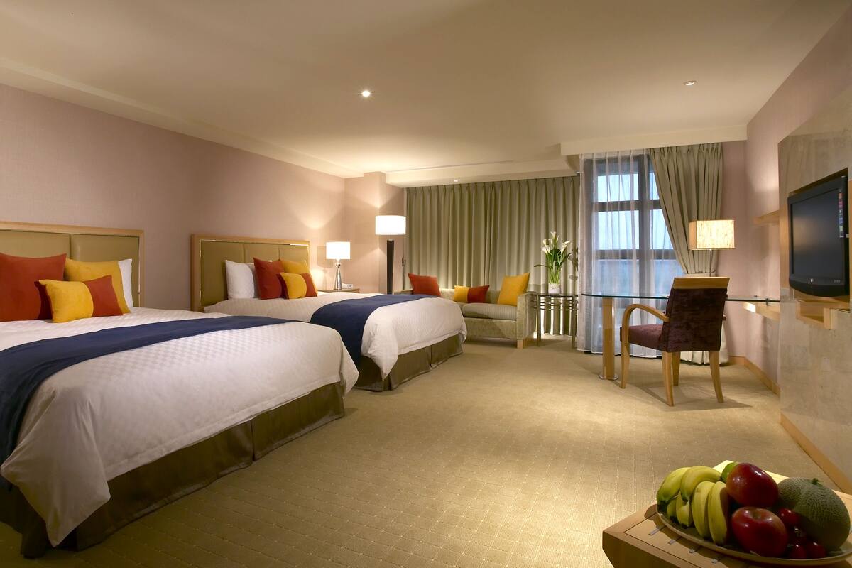 Quarto de hotel com duas camas, um sofá, uma televisão e uma cesta de frutas.