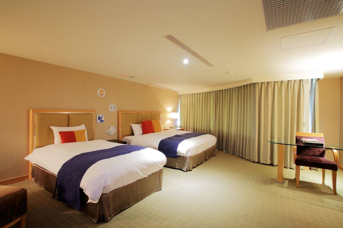Quarto de hotel com duas camas, uma mesa de vidro, uma cadeira e uma janela grande com cortinas.