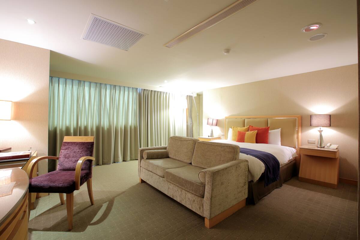 Quarto de hotel com cama, sofá, poltrona, escrivaninha e luminária.