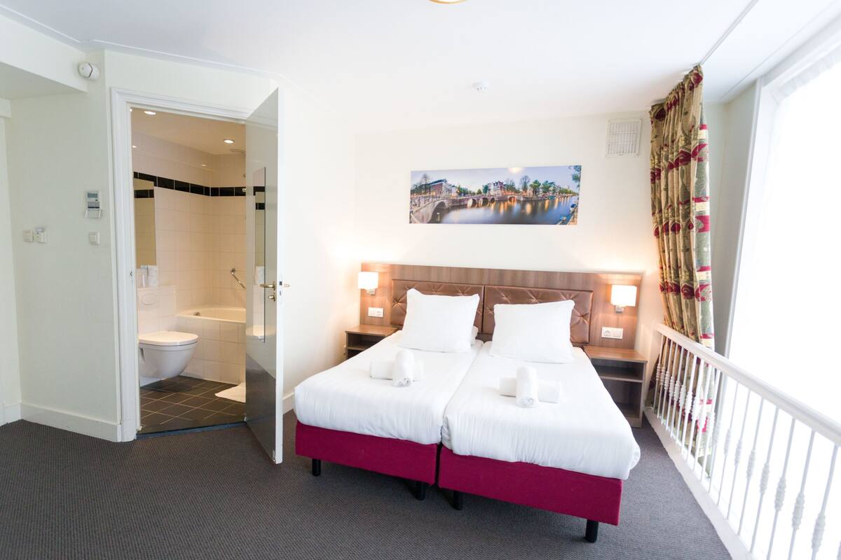 Quarto de hotel com cama de casal, banheiro com vaso sanitário e banheira, e varanda com grade.