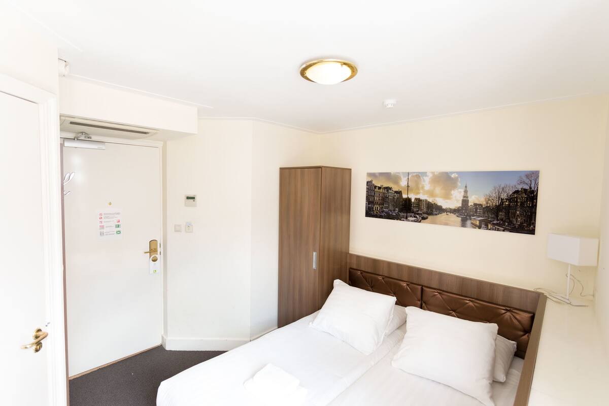 Quarto de hotel com uma cama, um guarda-roupa, uma porta com um sinal e uma pintura de paisagem urbana na parede.