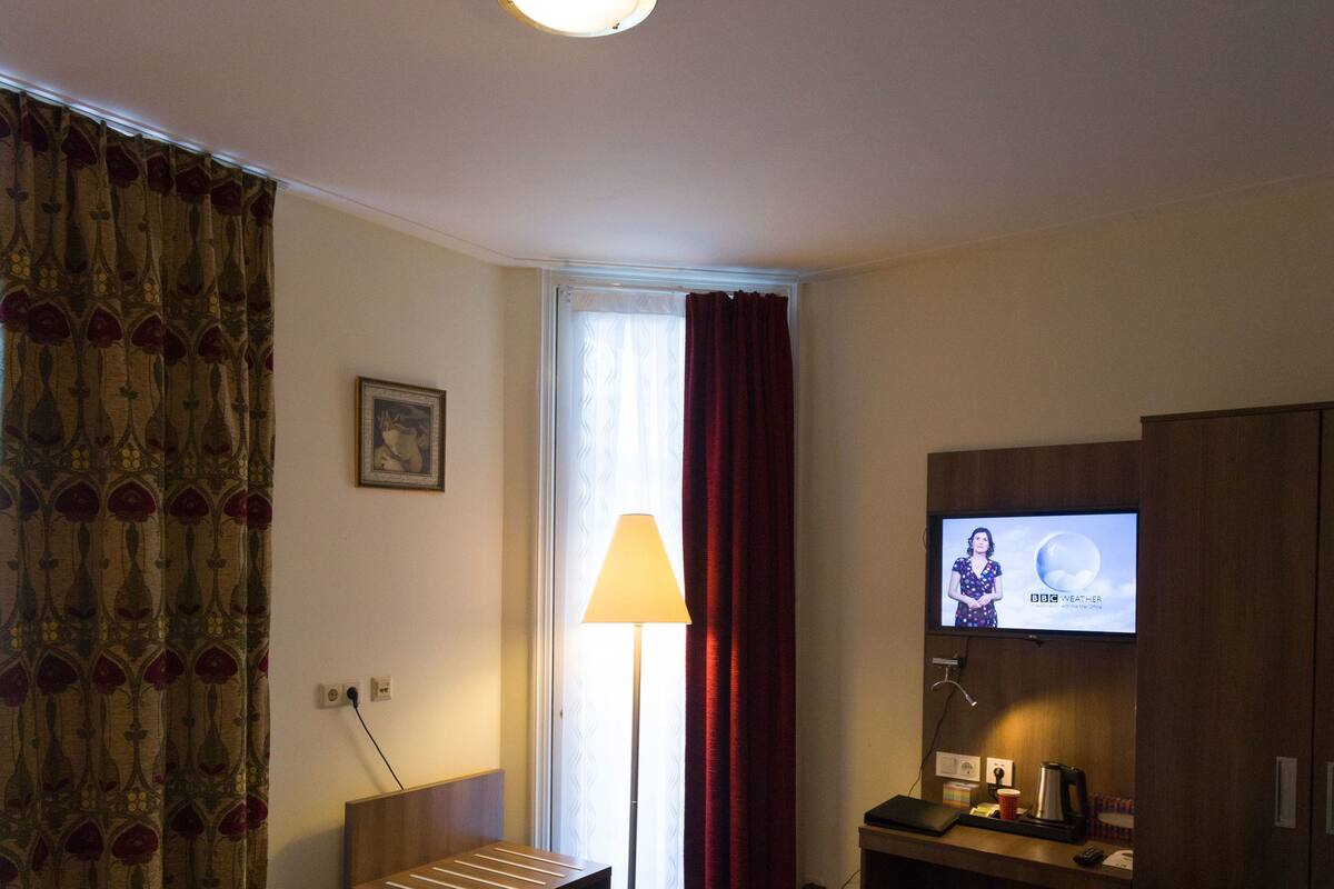 Quarto de hotel com cama, escrivaninha, televisão e janela com cortinas.