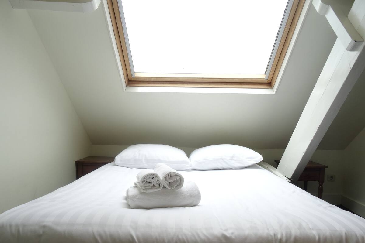 Cama bem arrumada em quarto com claraboia, roupa de cama branca e uma pequena mesa de cabeceira de madeira.