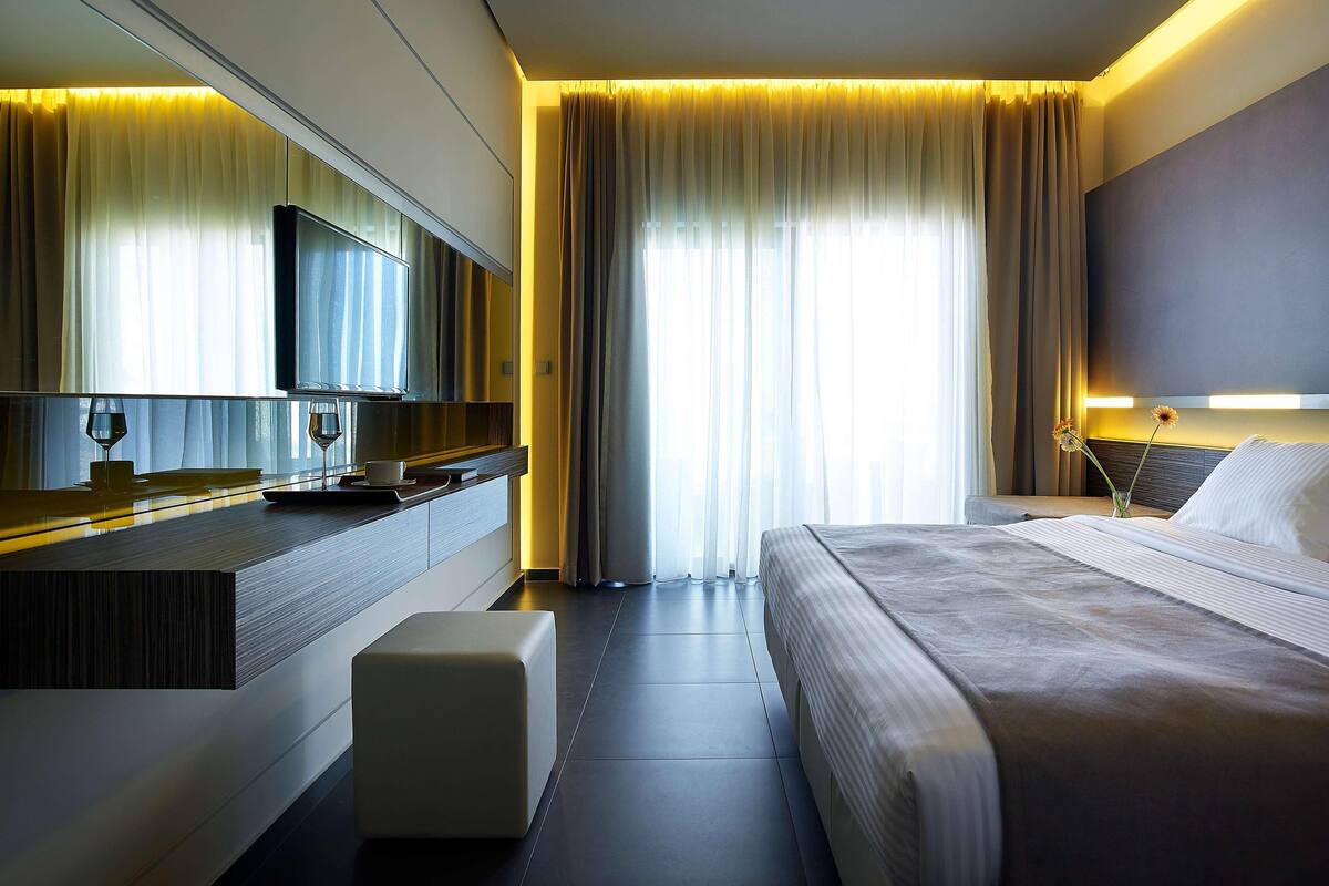 Quarto de hotel moderno com uma cama grande, mesa de cabeceira com abajur, televisão de tela plana e uma janela ampla com cortinas.