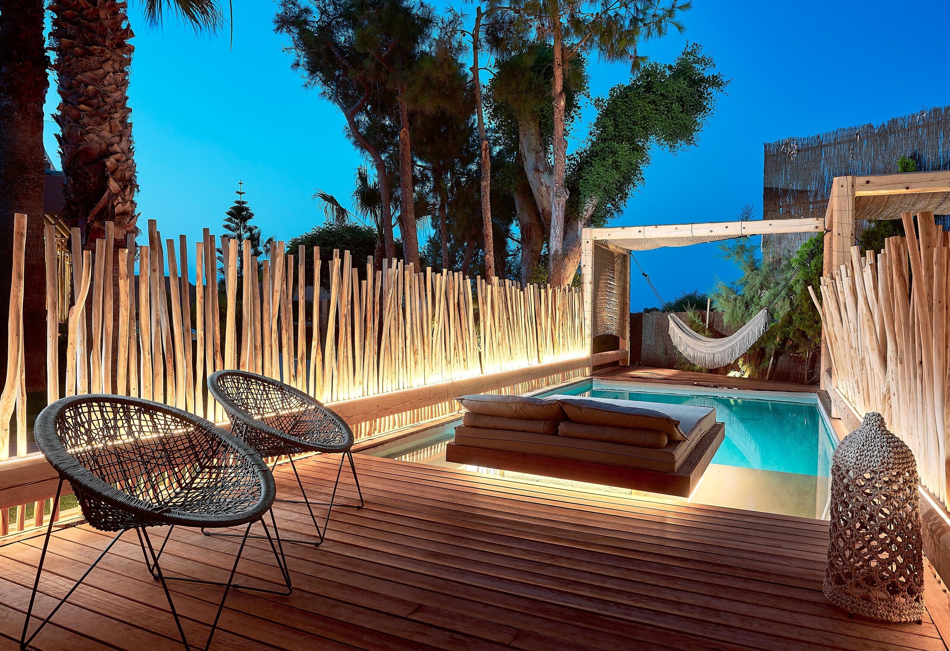 Um deck de madeira com piscina, duas cadeiras de vime, uma rede e um cercado de madeira.