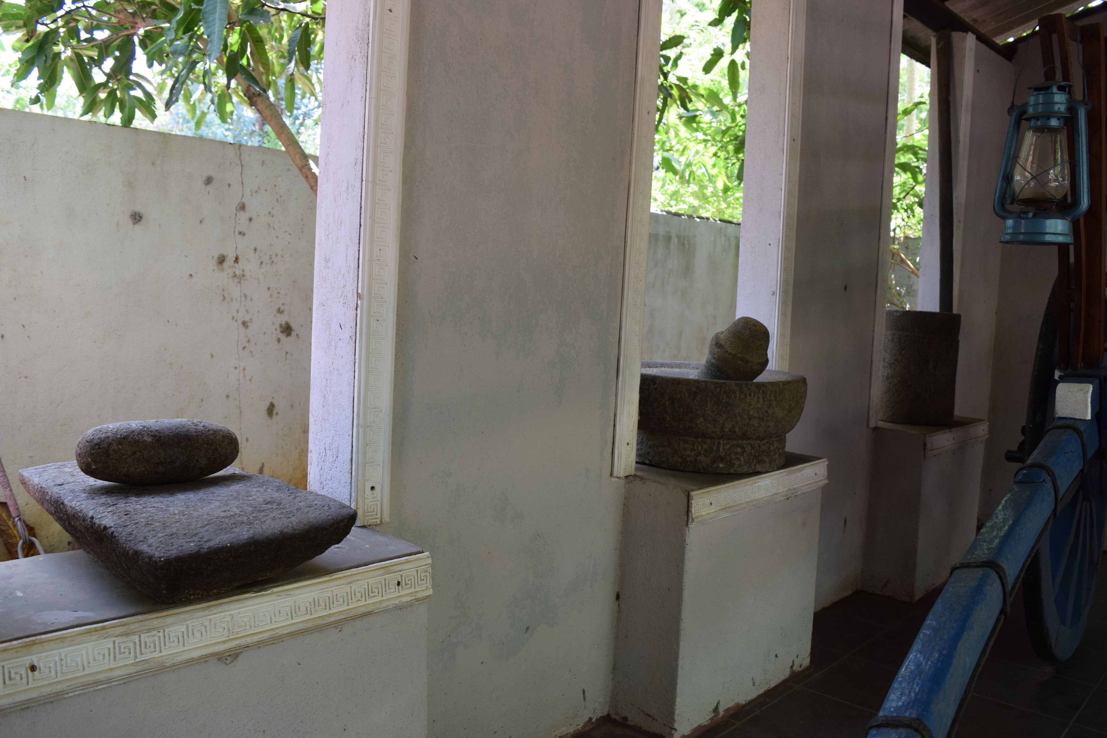 Um banco de pedra com almofada, uma bacia de pedra e um candeeiro num espaço com paredes brancas e uma janela.