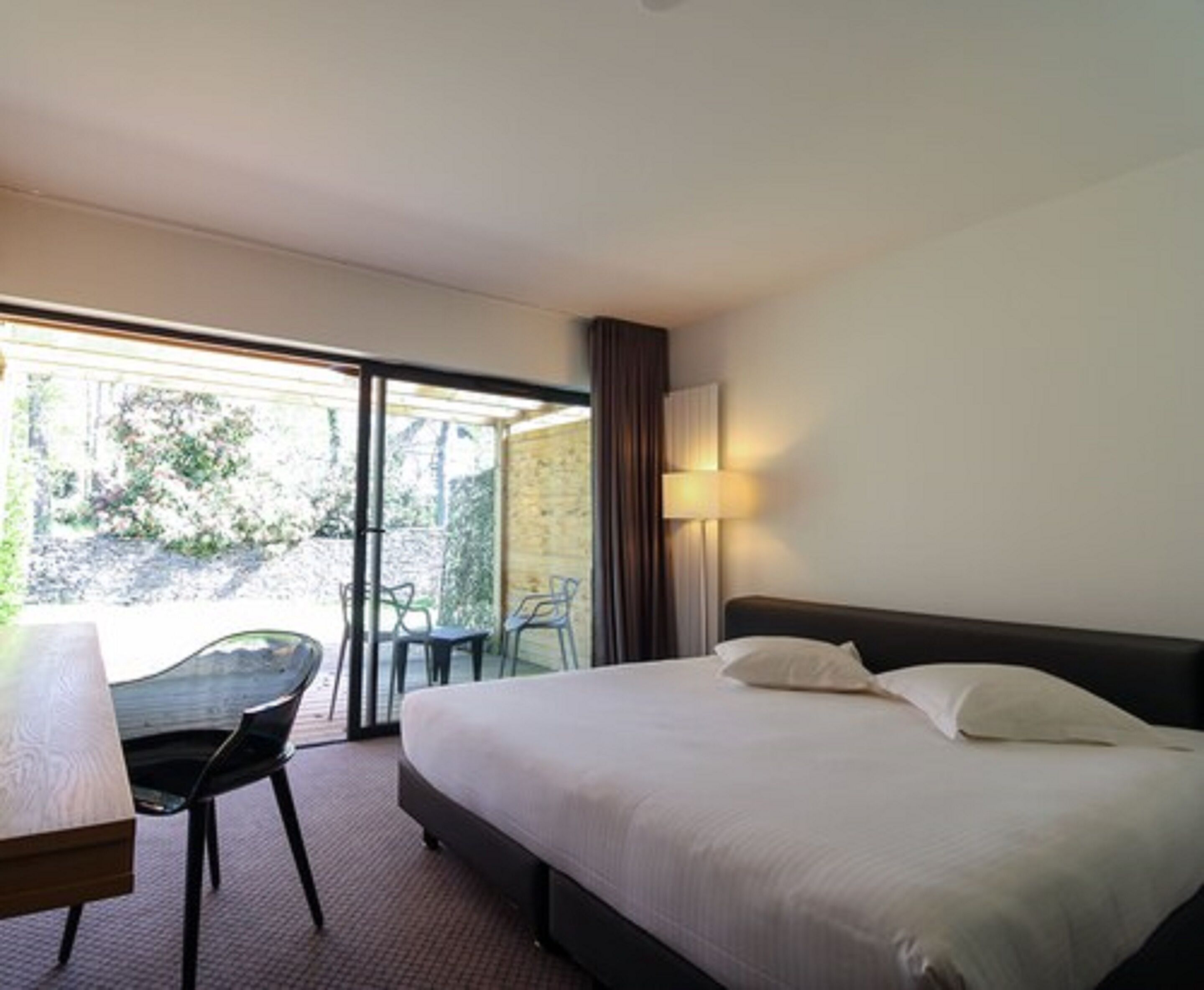 Quarto de hotel com cama, escrivaninha e cadeira. Porta de vidro deslizante dá acesso a área externa com cadeiras.