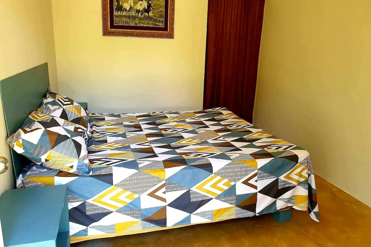 Uma cama de solteiro com colcha estampada, um quadro na parede e uma janela com cortina.