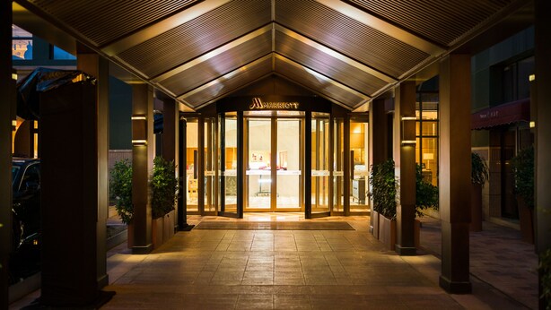 Entrada de hotel com design moderno, destacando um letreiro iluminado acima das portas e uma cobertura que leva à entrada.