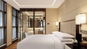 Quarto de hotel moderno com uma cama grande, uma mesa de cabeceira com abajur, uma pequena mesa com um vaso de flores e vista para o banheiro por uma porta de vidro.