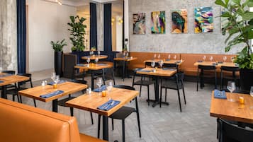 Um restaurante moderno com mesas de madeira e assentos laranja, apresentando arte abstrata nas paredes e plantas em vasos.
