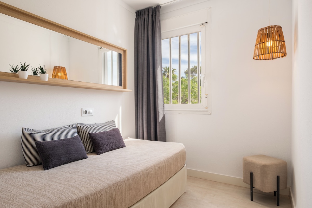 Quarto com uma cama, um banquinho pequeno, uma luminária pendente, uma janela com cortinas e uma estante com plantas.