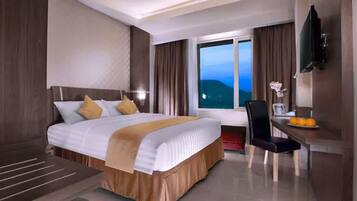 Quarto de hotel com uma cama grande, uma mesa com cadeira, televisão e janela com vista para a cidade ao entardecer.