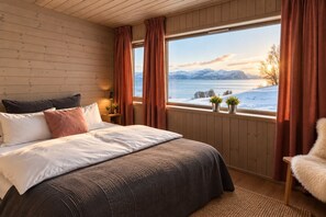 Quarto com uma janela grande, paredes de madeira, cama com várias almofadas, mesa de cabeceira e uma cadeira.