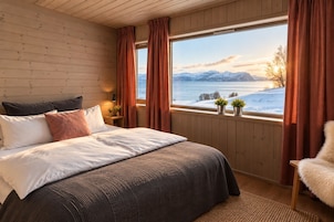 Quarto com uma janela grande, paredes de madeira, cama com várias almofadas, mesa de cabeceira e uma cadeira.