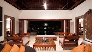 Um espaço amplo e interno, com mobiliário de madeira, uma mesa central e vista para um recurso hídrico externo.
