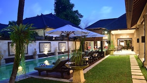 Área da piscina com espreguiçadeiras e guarda-sóis, um gramado bem cuidado e um edifício com telhado em azulejo.