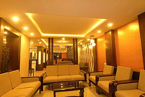 Um saguão de hotel com sofás, cadeiras, uma mesa de centro de vidro e uma área de recepção ao fundo.
