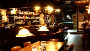 Uma área de bar com mesas e cadeiras de madeira, um balcão de recepção e prateleiras com garrafas e copos.