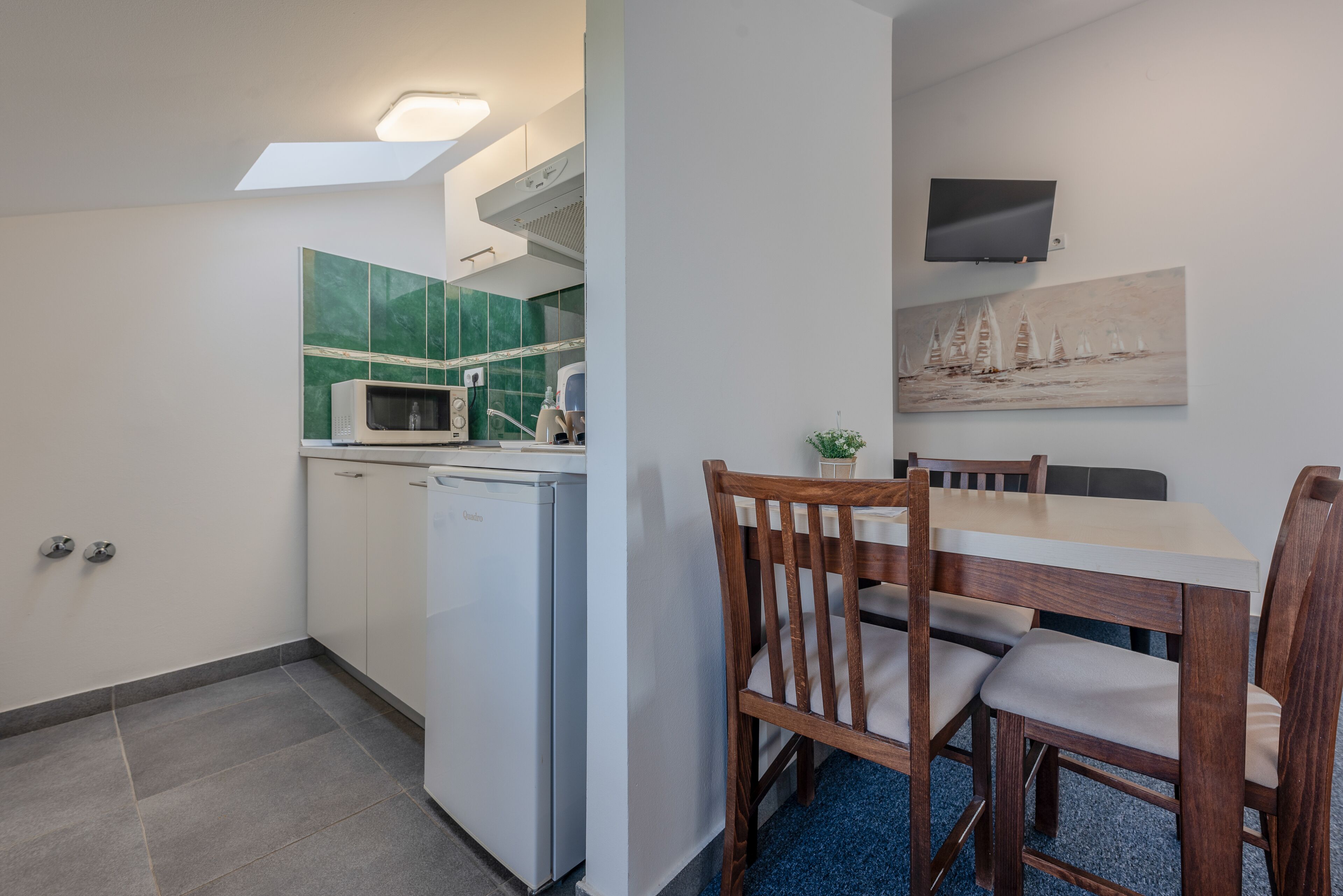 Uma cozinha compacta com micro-ondas, mini-geladeira e painel atrás do fogão verde. Há uma mesa de jantar com cadeiras de madeira e uma TV de tela plana presa à parede.