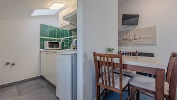 Uma cozinha compacta com micro-ondas, mini-geladeira e painel atrás do fogão verde. Há uma mesa de jantar com cadeiras de madeira e uma TV de tela plana presa à parede.